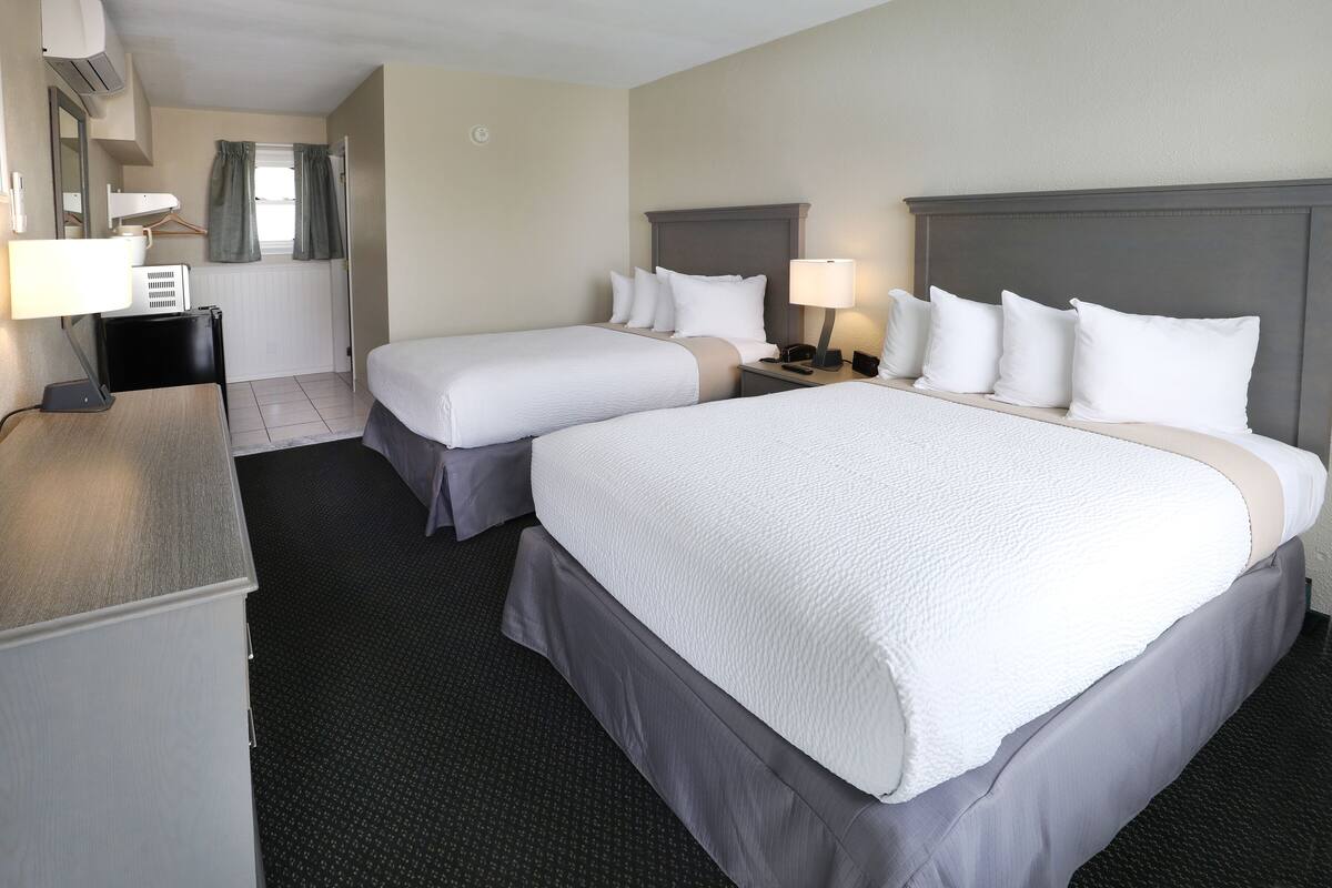 Quarto de hotel com duas camas, uma mesa, uma cadeira, um abajur e uma janela com cortinas.