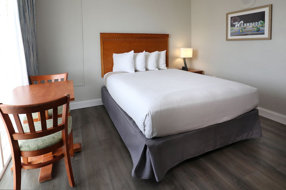 Cama bem arrumada com cabeceira de madeira, mesa de cabeceira pequena com abajur e cadeira com almofada.