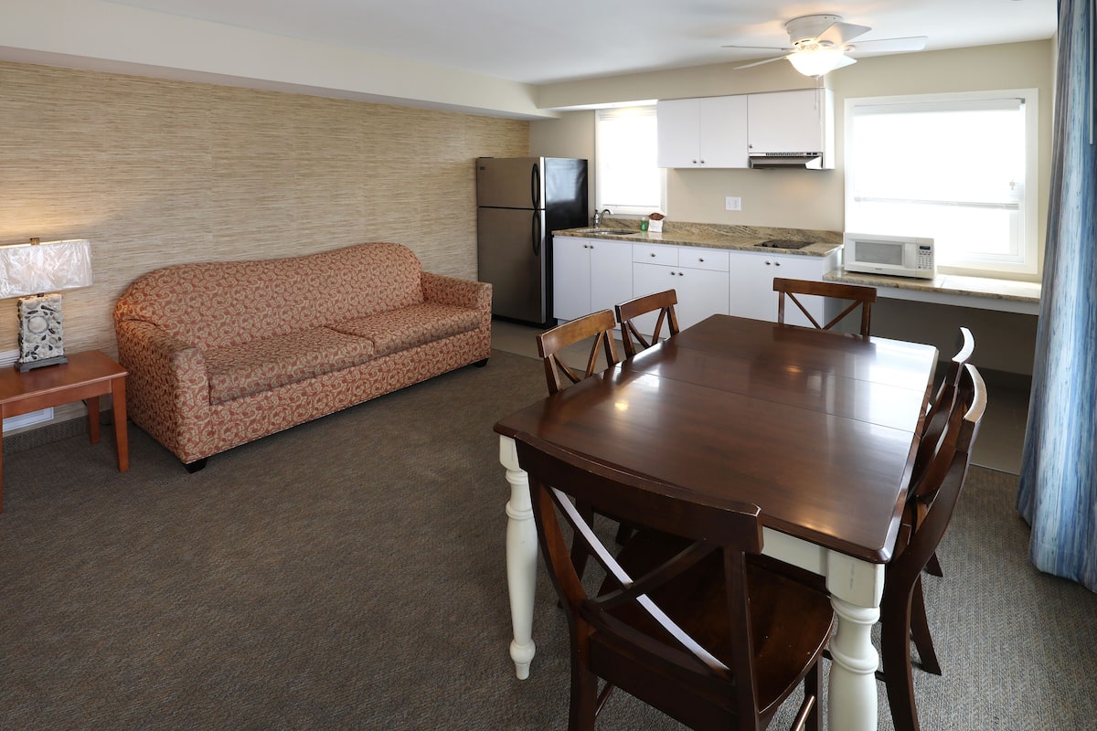 Um espaço compacto com sofá, mesa de jantar, cadeiras, frigorífico, micro-ondas e janela com cortinas.