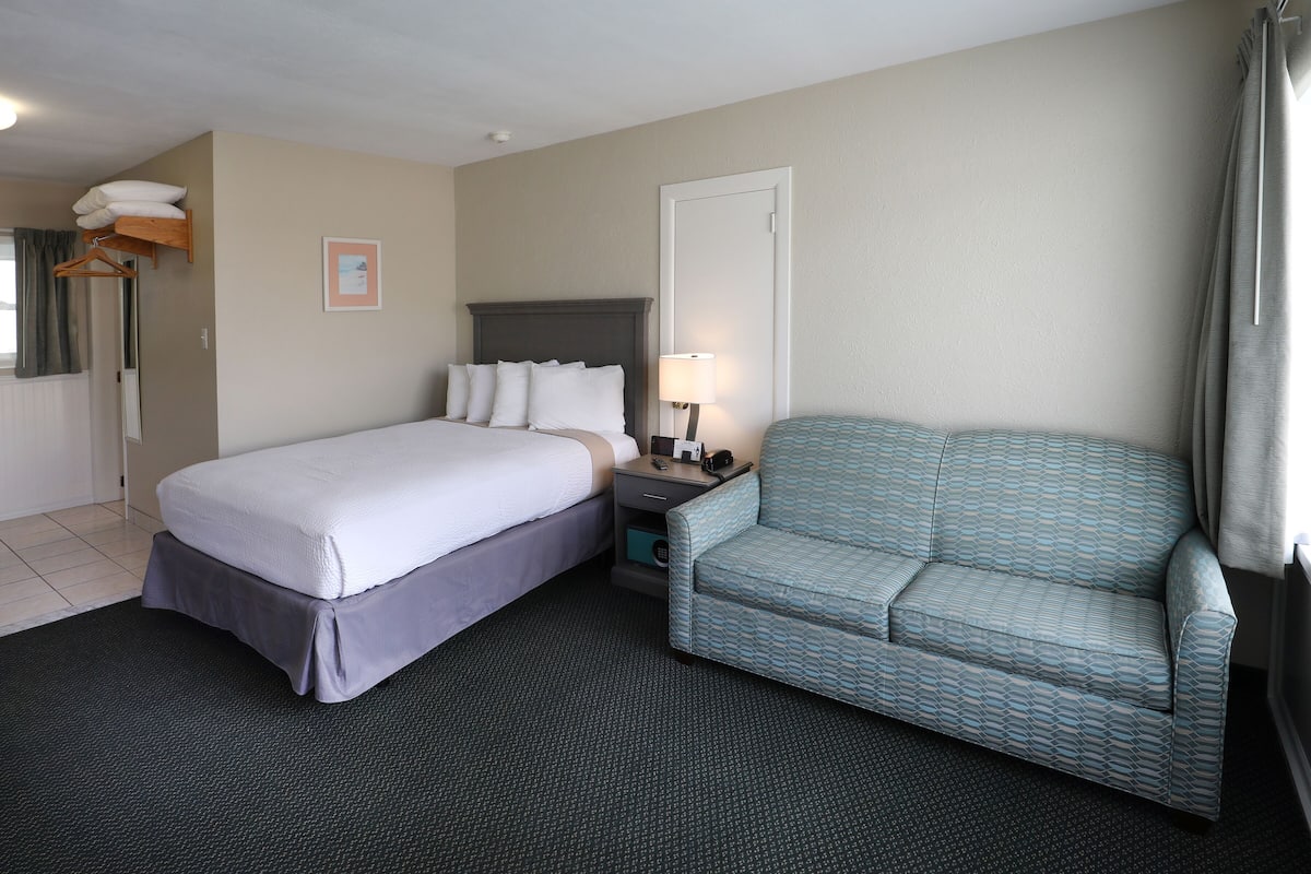 Quarto de hotel com cama, mesa de cabeceira, luminária, sofá e cabideiro.
