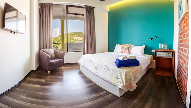 Quarto de hotel com cama, poltrona, televisão, janela com cortinas e vista para edifícios.