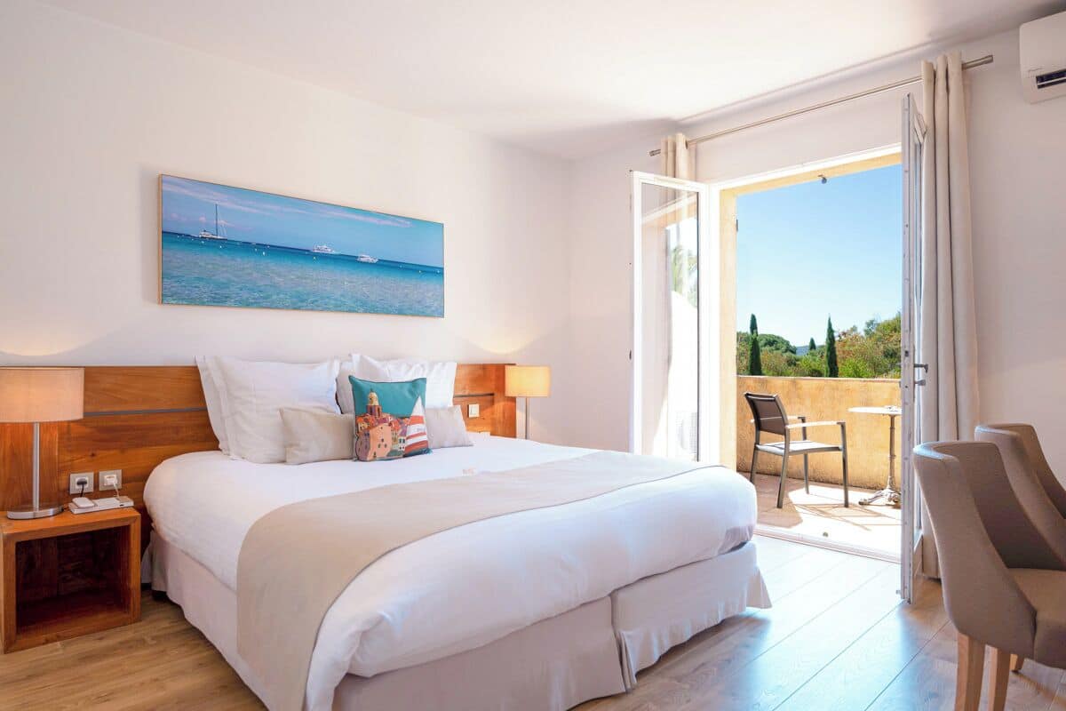 Quarto de hotel com uma cama grande, uma pintura de paisagem marítima, varanda com mesa e cadeiras, e vista para o mar.