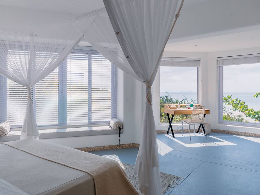 Um quarto com cama, escrivaninha e amplas janelas com vista para o mar.