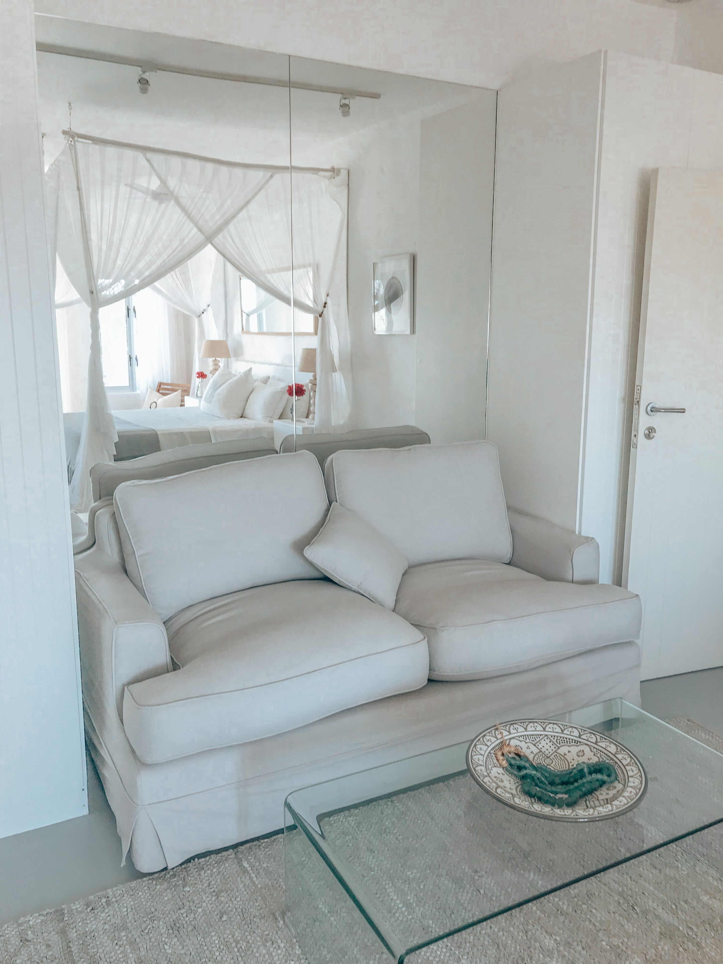 Uma sala de estar moderna e minimalista com um sofá bege, uma mesa de centro de vidro e uma cama com dossel ao fundo.