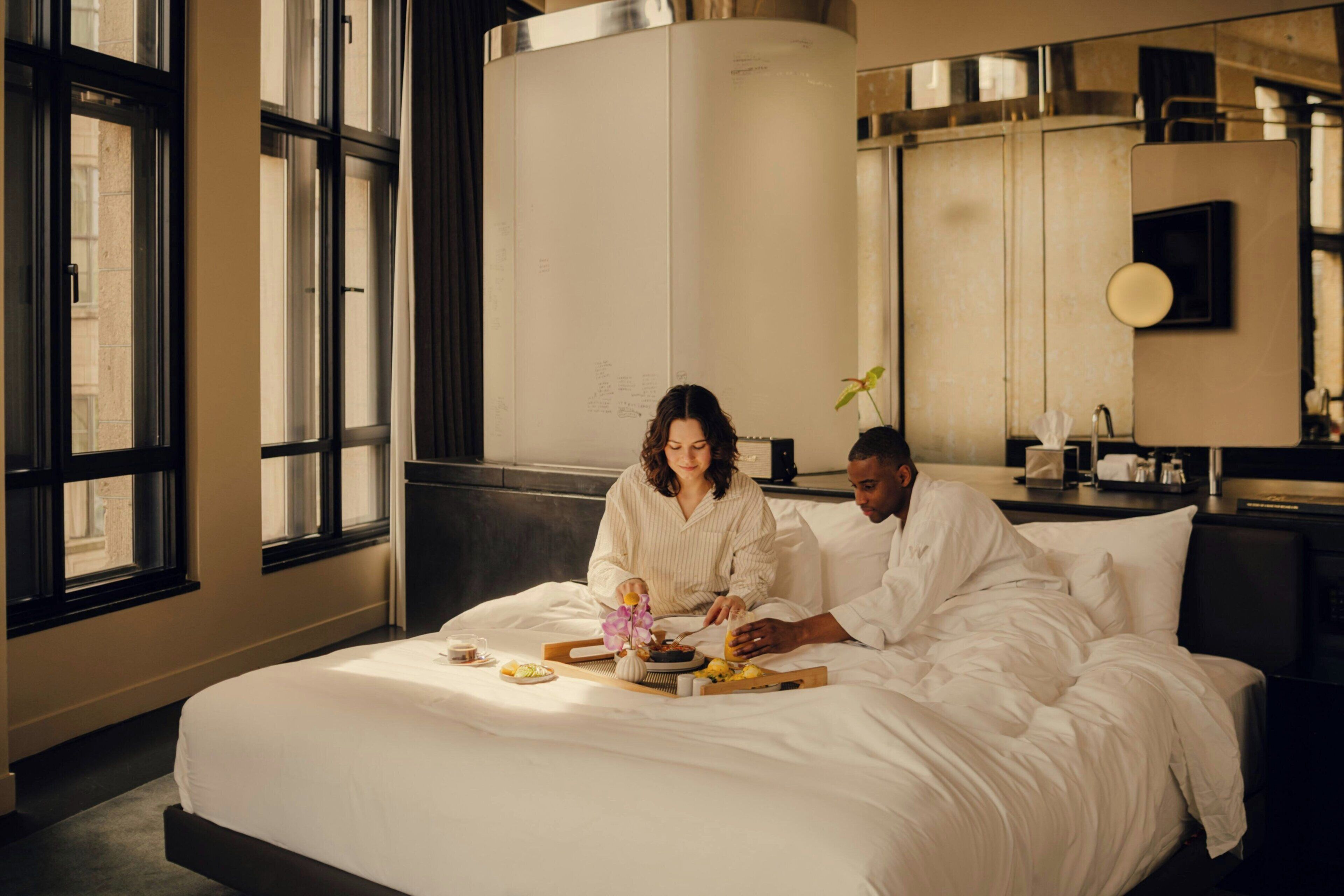 Quarto de hotel com uma cama grande, mesa de cabeceira com bandeja de comida, janela e espelho.