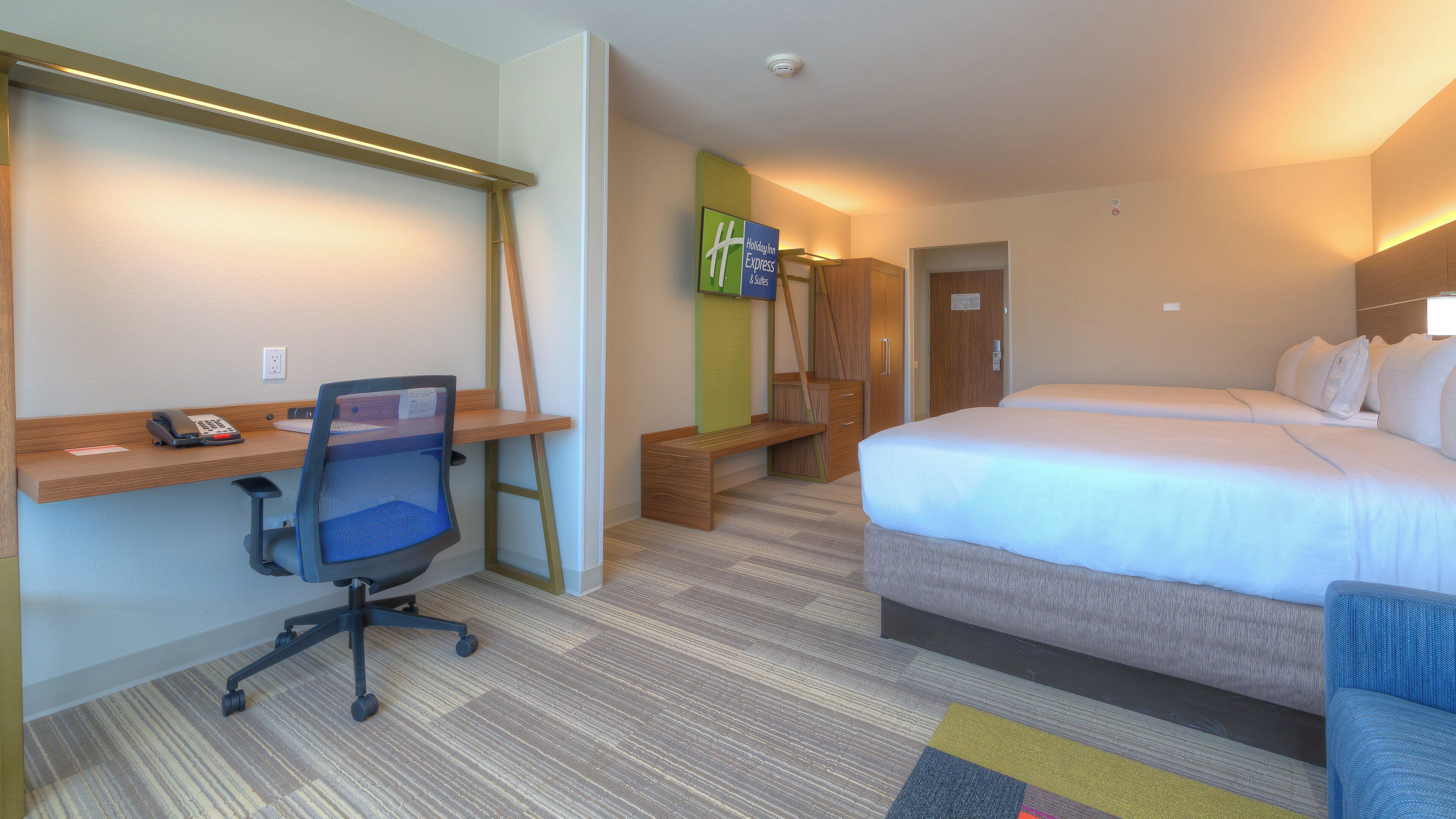 Quarto de hotel com escrivaninha, cadeira, cama e televisão na parede.