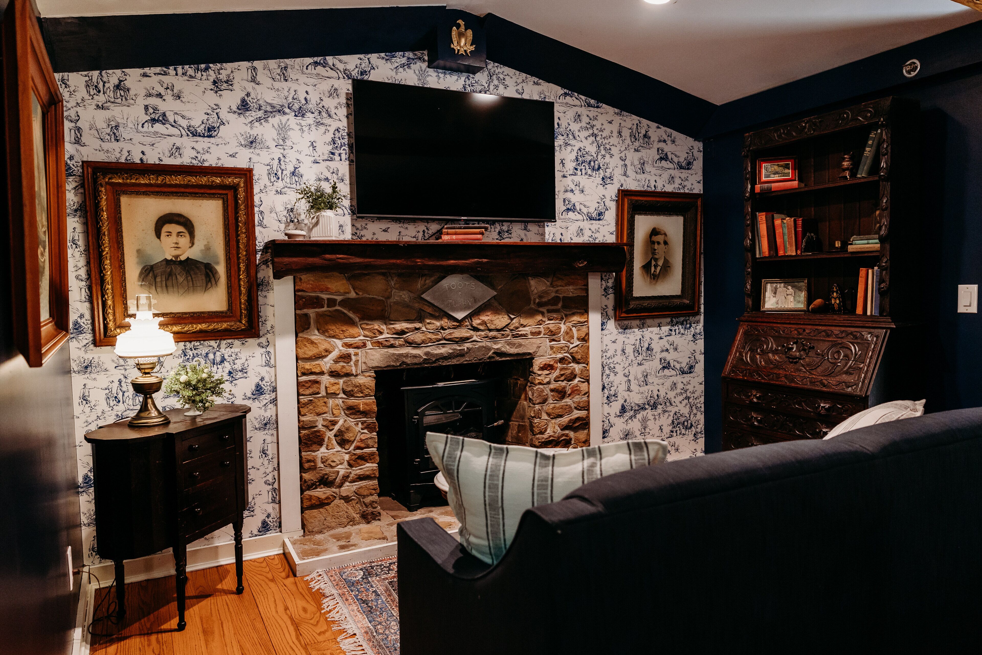 Uma sala de estar acolhedora, com lareira de pedra, televisão de tela plana acima e uma estante repleta de livros e objetos decorativos.