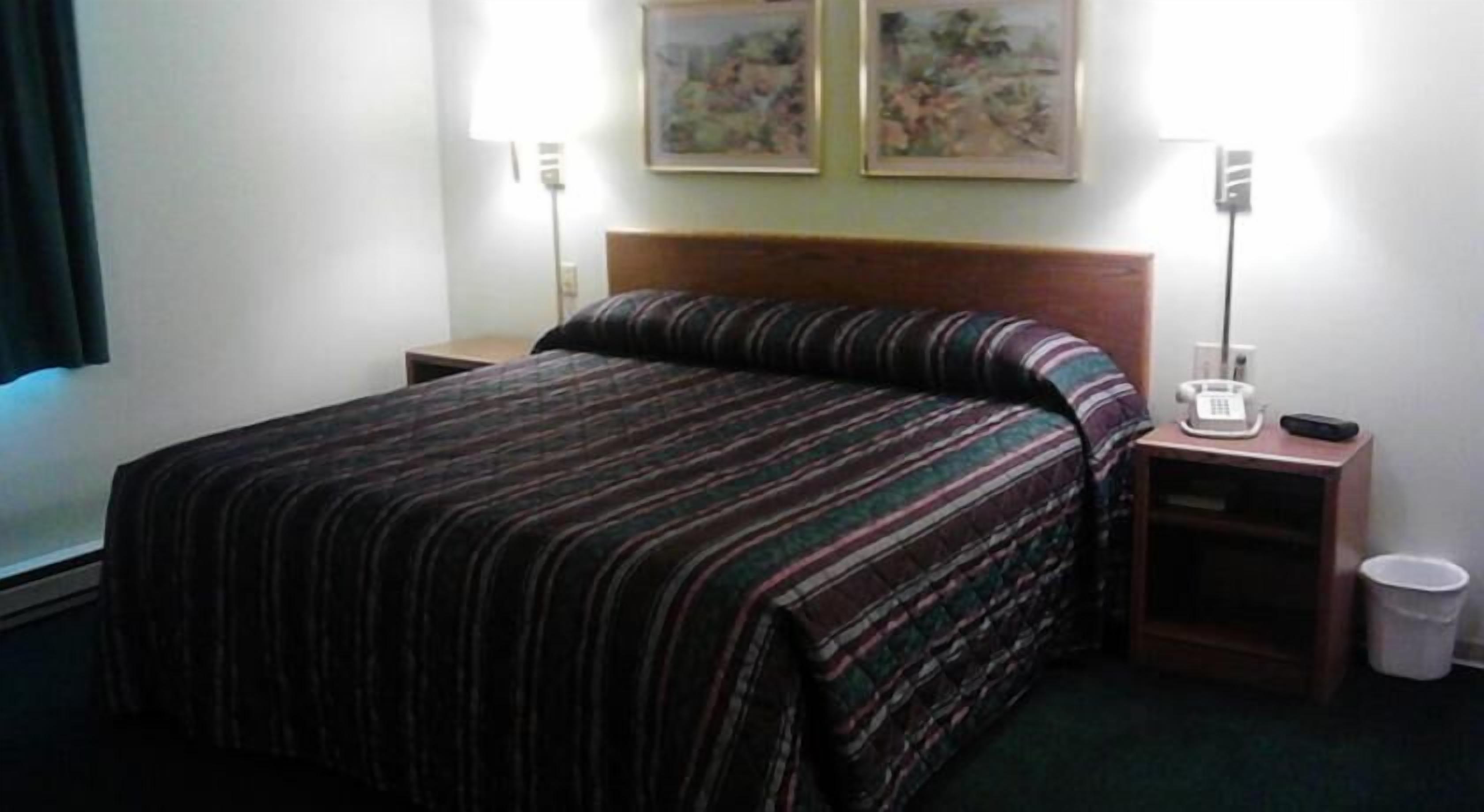 Uma cama bem arrumada com um cobertor listrado, uma mesinha de cabeceira com abajur e uma lixeira.
