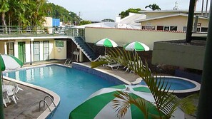 Piscina com espreguiçadeiras e guarda-sóis, uma escadaria e um prédio com telhado vermelho ao fundo.
