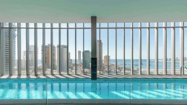Piscina interna moderna com vista para a cidade, janelas amplas e céu claro.
