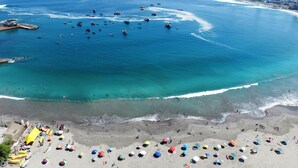 Vista aérea de uma praia com barcos, um píer e o horizonte da cidade ao fundo.