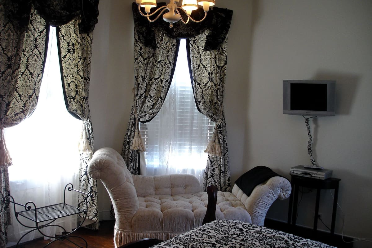 Quarto com uma cama grande, uma poltrona branca, uma mesinha com uma televisão e cortinas estampadas.