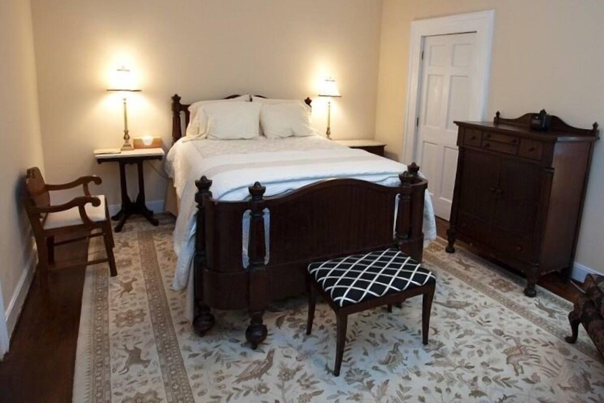 Quarto com cama, duas mesinhas de cabeceira, uma cadeira, cômoda e tapete estampado.