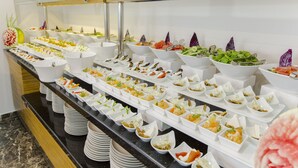 Buffet com diversos pratos dispostos em tigelas brancas sobre bandejas pretas, em frente a uma bancada de mármore e parede branca.