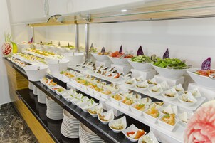 Buffet com diversos pratos dispostos em tigelas brancas sobre bandejas pretas, em frente a uma bancada de mármore e parede branca.