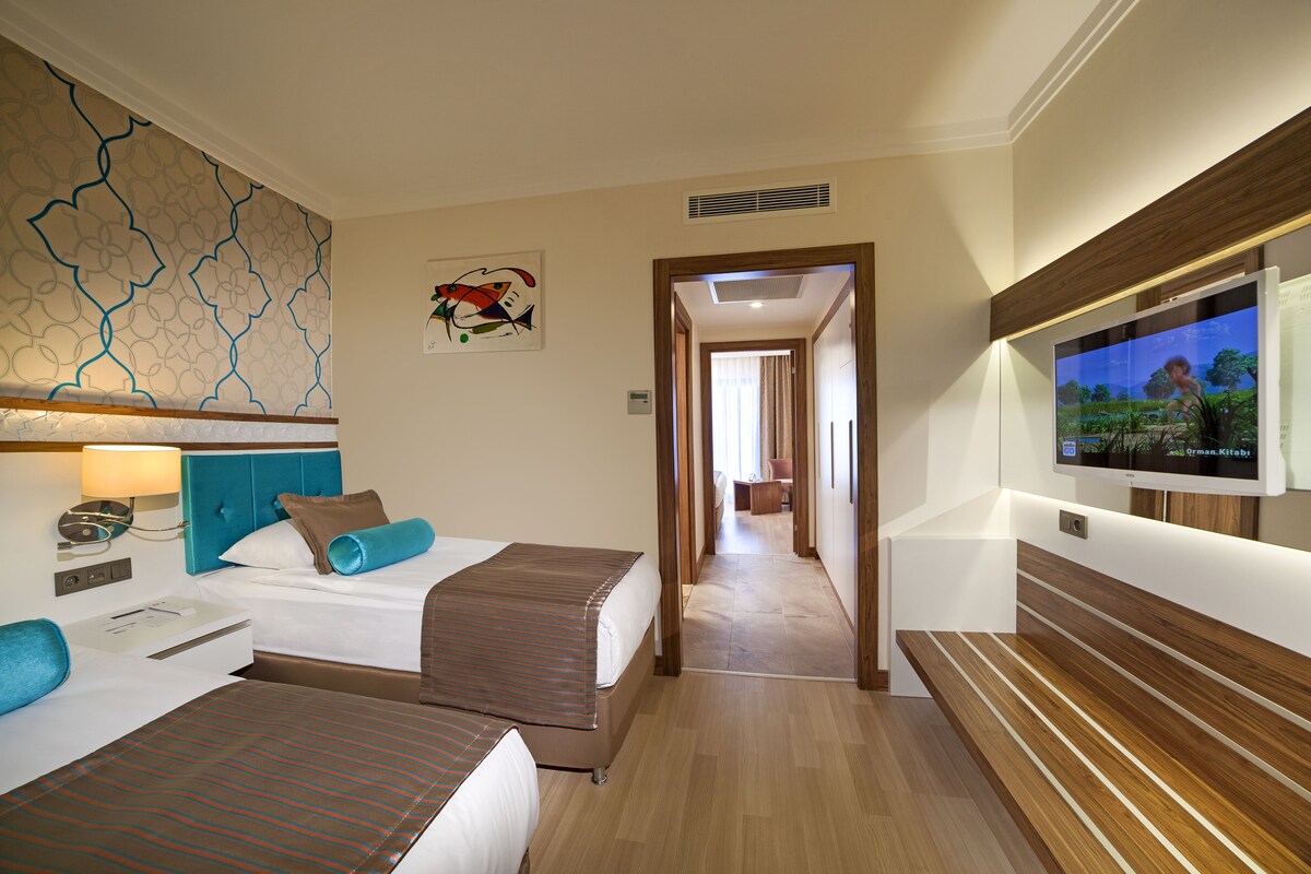 Quarto de hotel com duas camas, uma televisão e corredor que dá para outro quarto.
