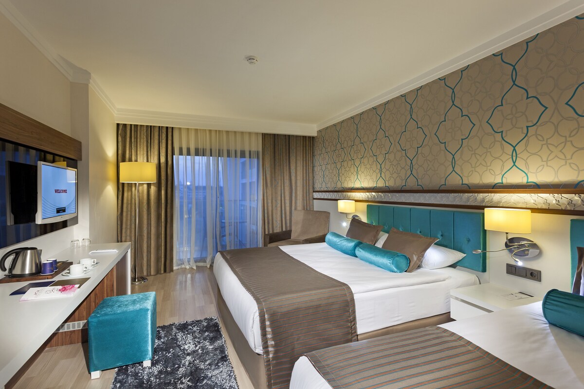 Quarto de hotel com uma cama grande, uma escrivaninha com computador, uma cadeira e televisão na parede.