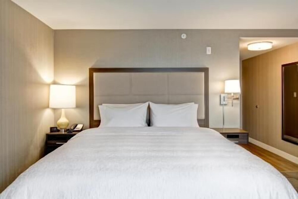 Quarto de hotel com uma cama grande, duas mesas de cabeceira com abajures, uma cabeceira de madeira e uma porta à direita.