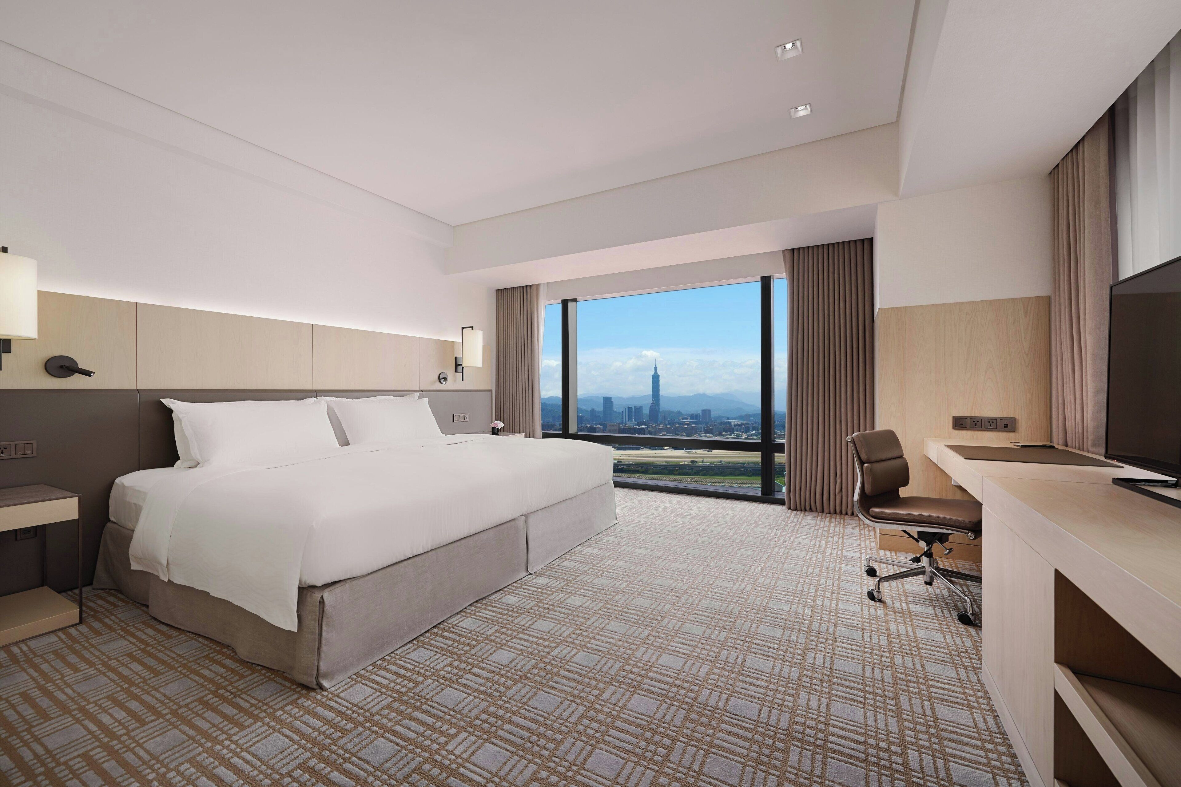 Quarto de hotel com uma cama grande, uma escrivaninha, uma cadeira e uma televisão.
