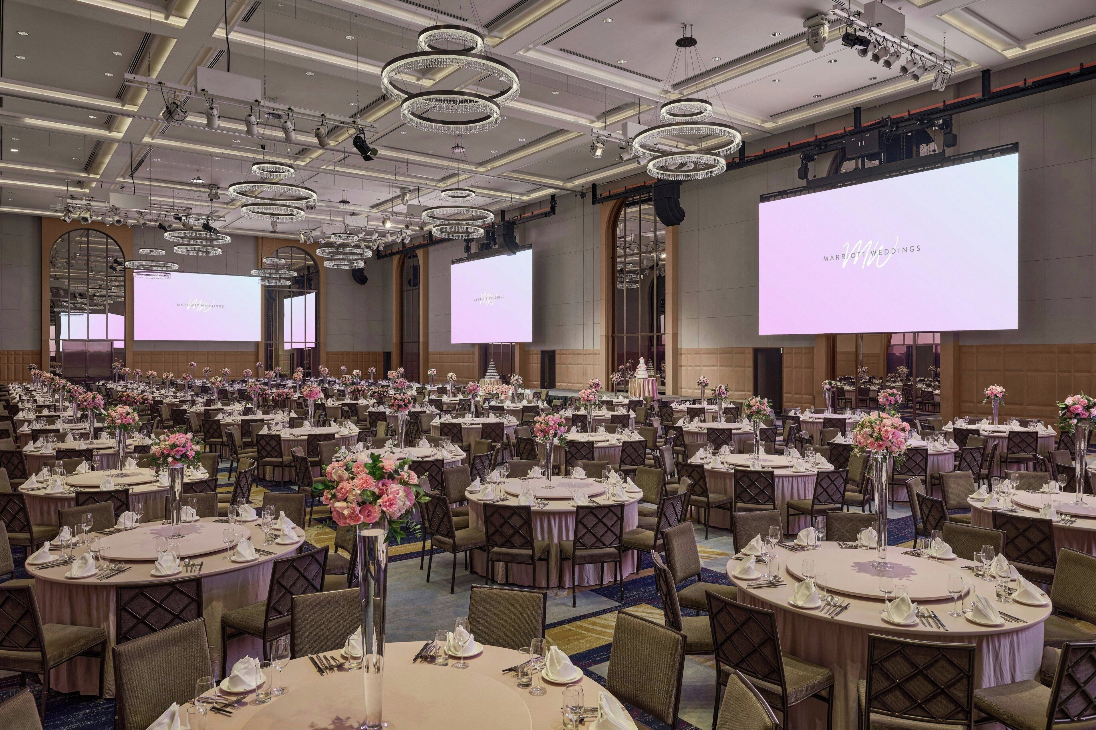 Um amplo salão de banquetes com mesas redondas preparadas para um casamento, decorado com arranjos florais e toalhas brancas.