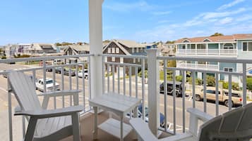 Varanda com cadeiras Adirondack brancas e uma pequena mesa, com vista para uma rua com carros estacionados e casas.