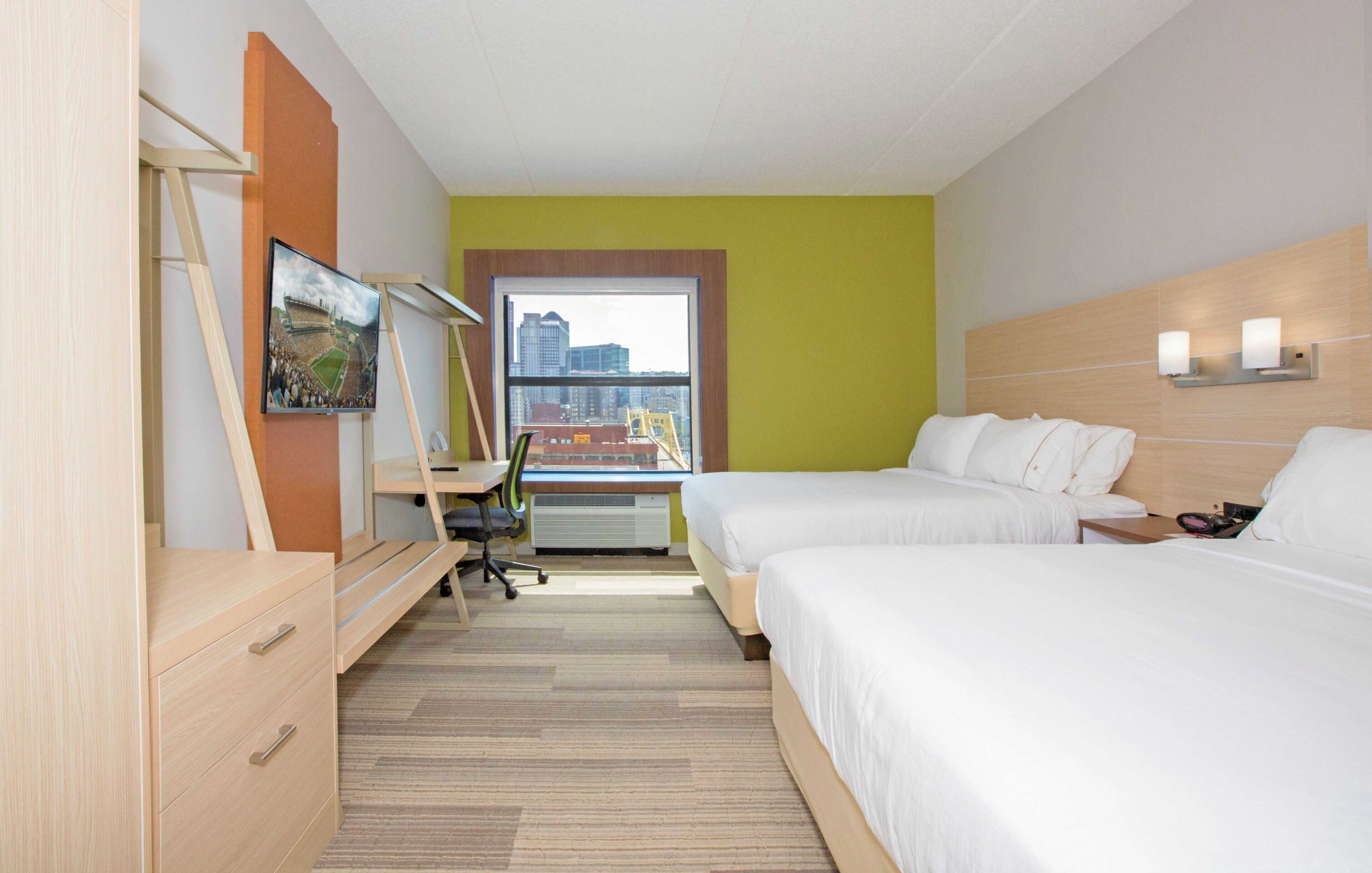 Quarto de hotel com duas camas, uma escrivaninha com cadeira, televisão na parede e janela com vista para a cidade.