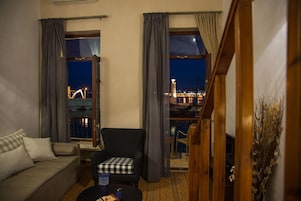 Sala de estar com sofá, poltrona e mesa de centro, janelas amplas com cortinas e vista para a cidade iluminada à noite.