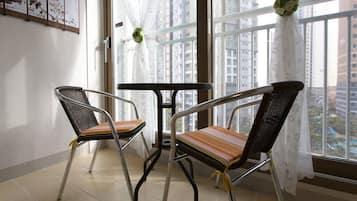 Quarto com duas cadeiras, uma mesa pequena, janelas amplas com cortinas e vista para edifícios altos.