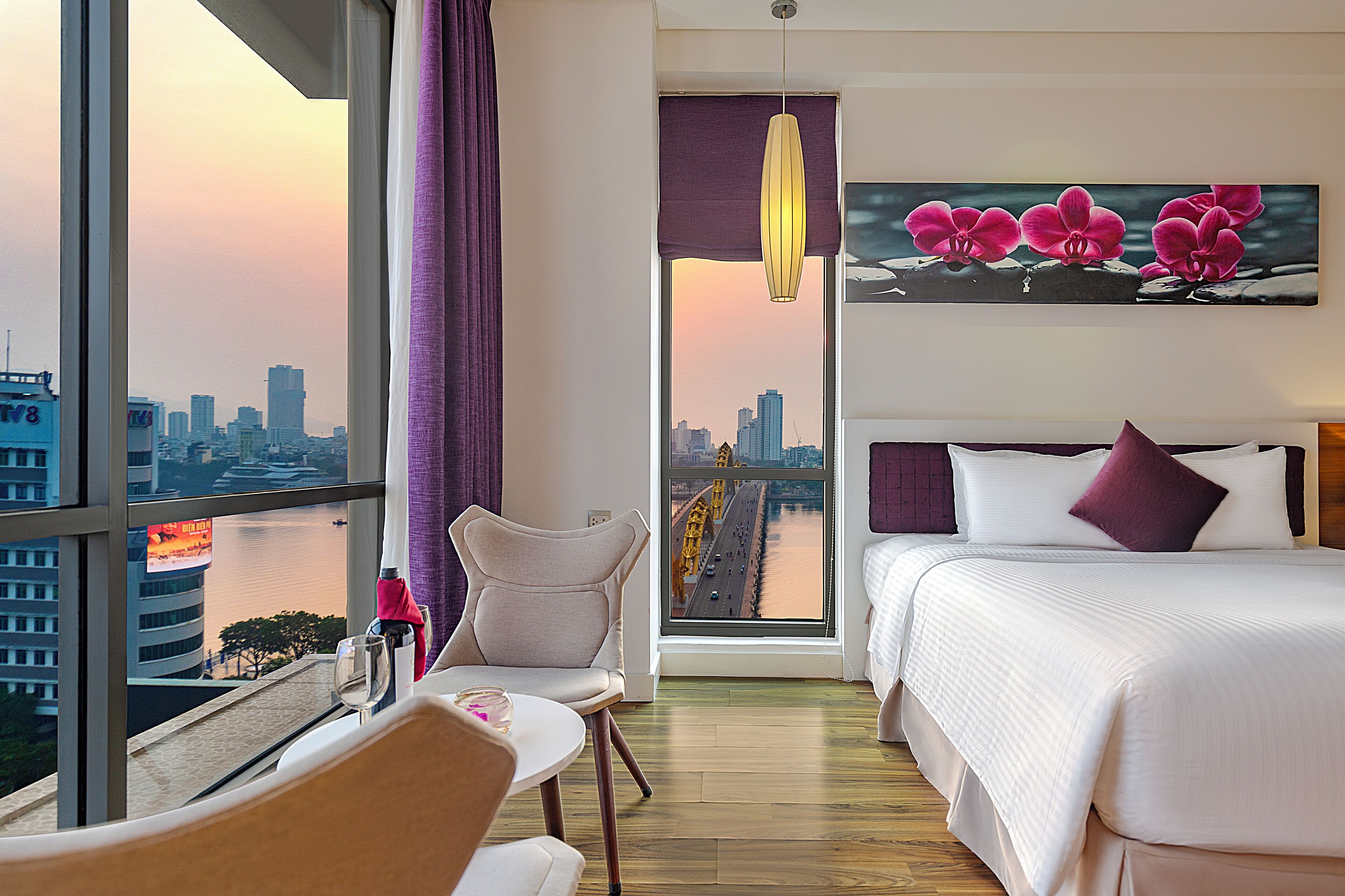 Quarto de hotel com uma grande janela, uma cama, uma cadeira e um quadro com flores cor-de-rosa.