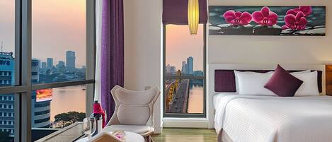 Quarto de hotel com uma grande janela, uma cama, uma cadeira e um quadro com flores cor-de-rosa.