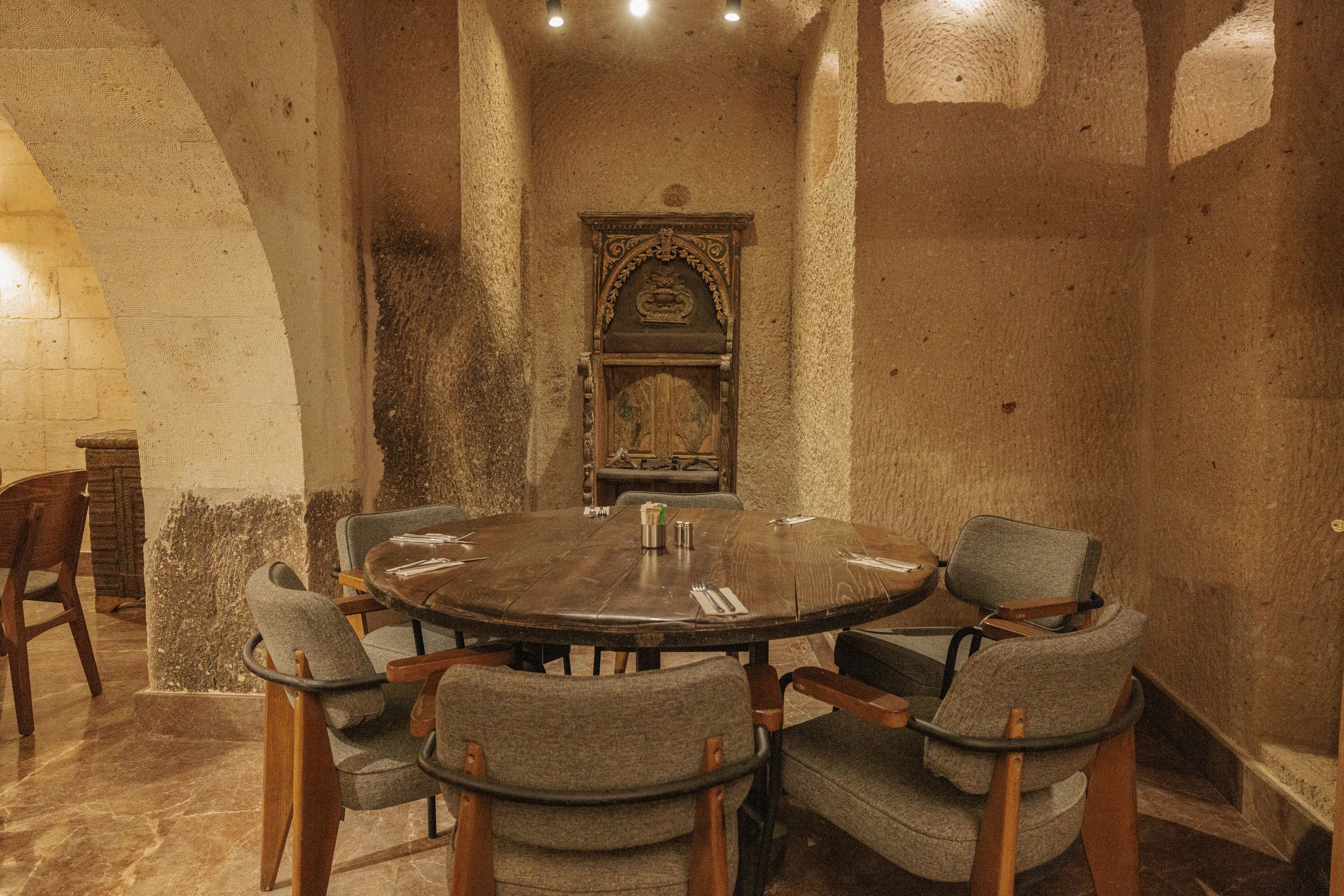 Uma mesa redonda de madeira com seis cadeiras, um armário de madeira com topo decorativo e uma parede de pedra com uma porta em arco.