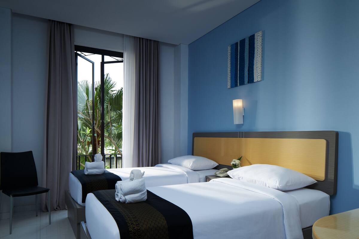 Quarto de hotel com duas camas, uma cadeira, uma escrivaninha e vista para palmeiras.