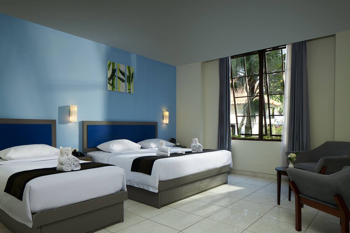 Quarto de hotel com duas camas, área de estar e uma grande janela vista para palmeiras.