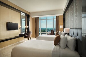 Quarto de hotel com duas camas, uma mesa, televisão e uma grande janela com vista para a cidade.