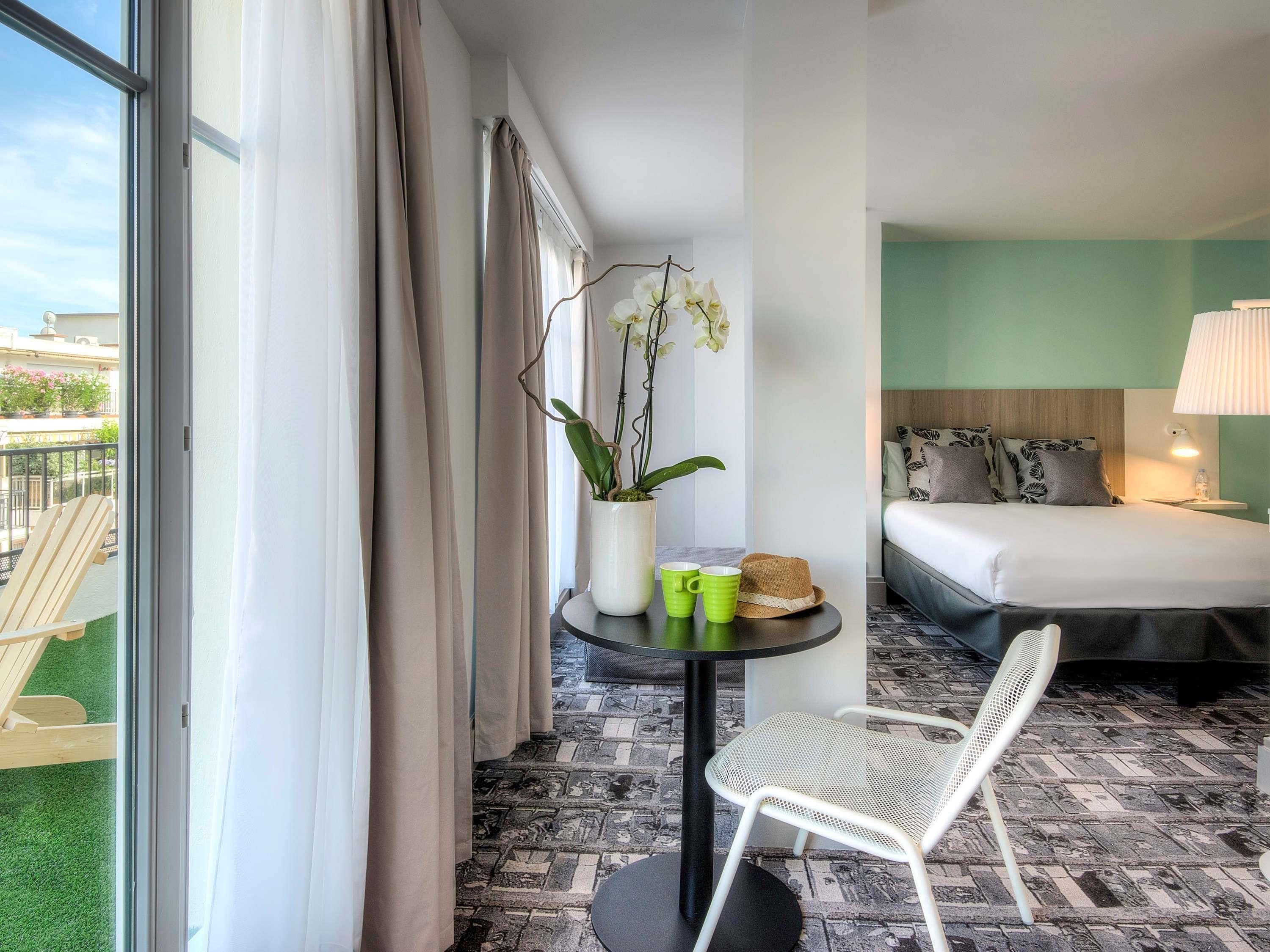 Quarto de hotel com cama, poltrona, mesa e uma orquídea em vaso, e vista para a varanda externa.