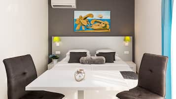 Quarto de hotel moderno com uma cama branca, duas cadeiras escuras, um quadro na parede e uma mesinha com uma lâmpada e um frasco.