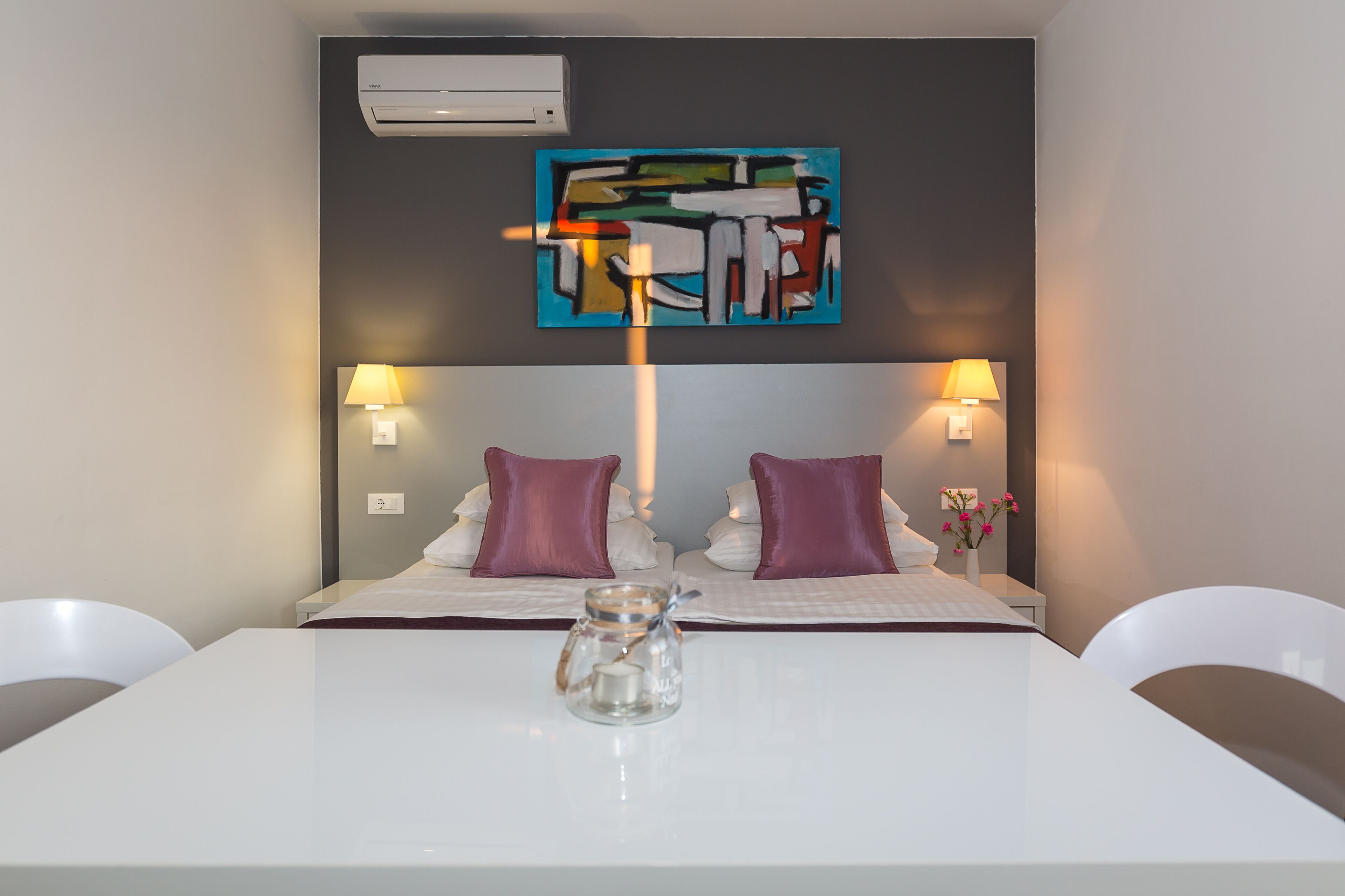 Quarto de hotel moderno com uma cama, duas luminárias de cabeceira, uma mesinha com um vaso e um quadro na parede.