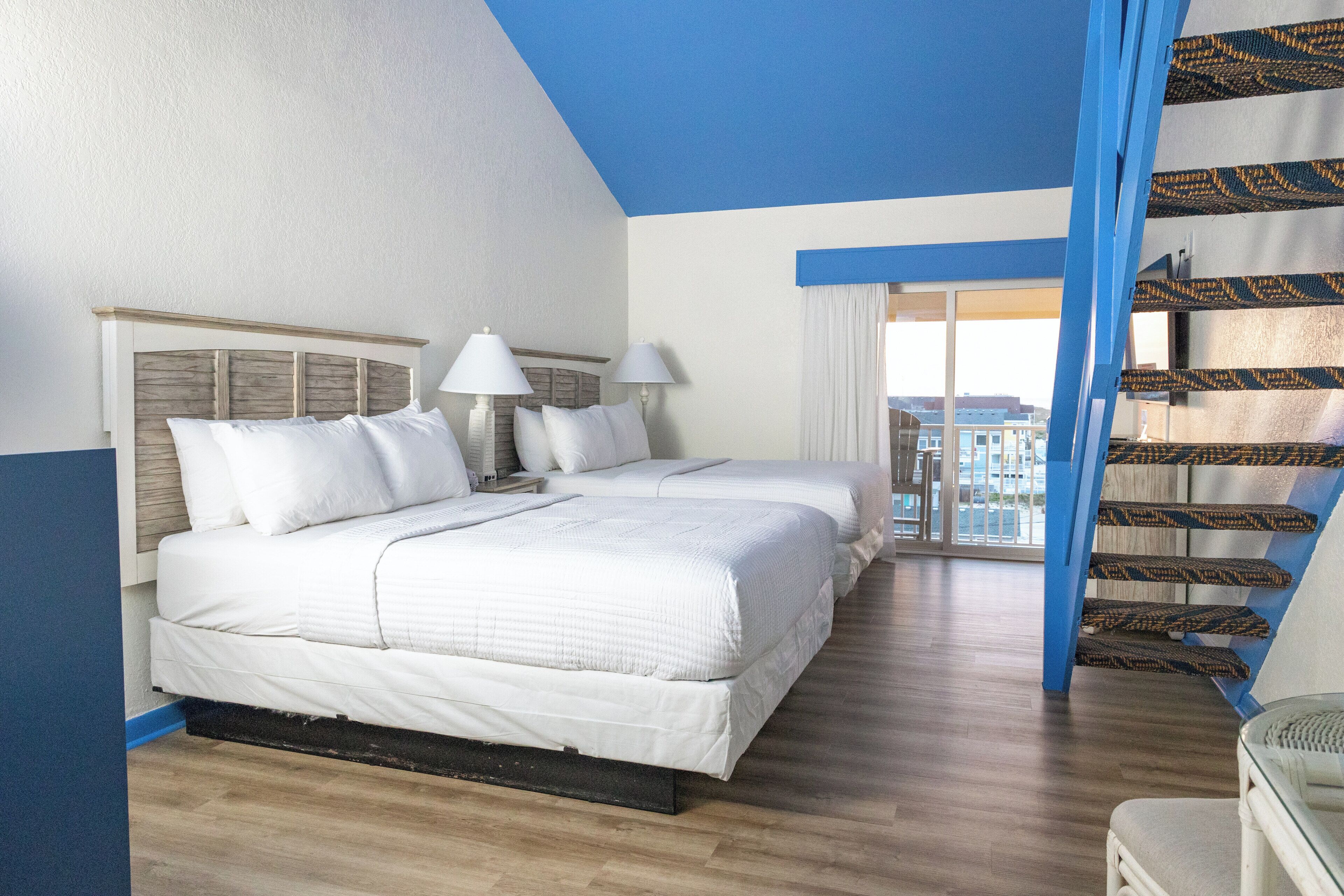 Quarto de hotel com duas camas, cabeceira de madeira, roupa de cama branca, uma janela grande com cortinas e uma escadaria azul.