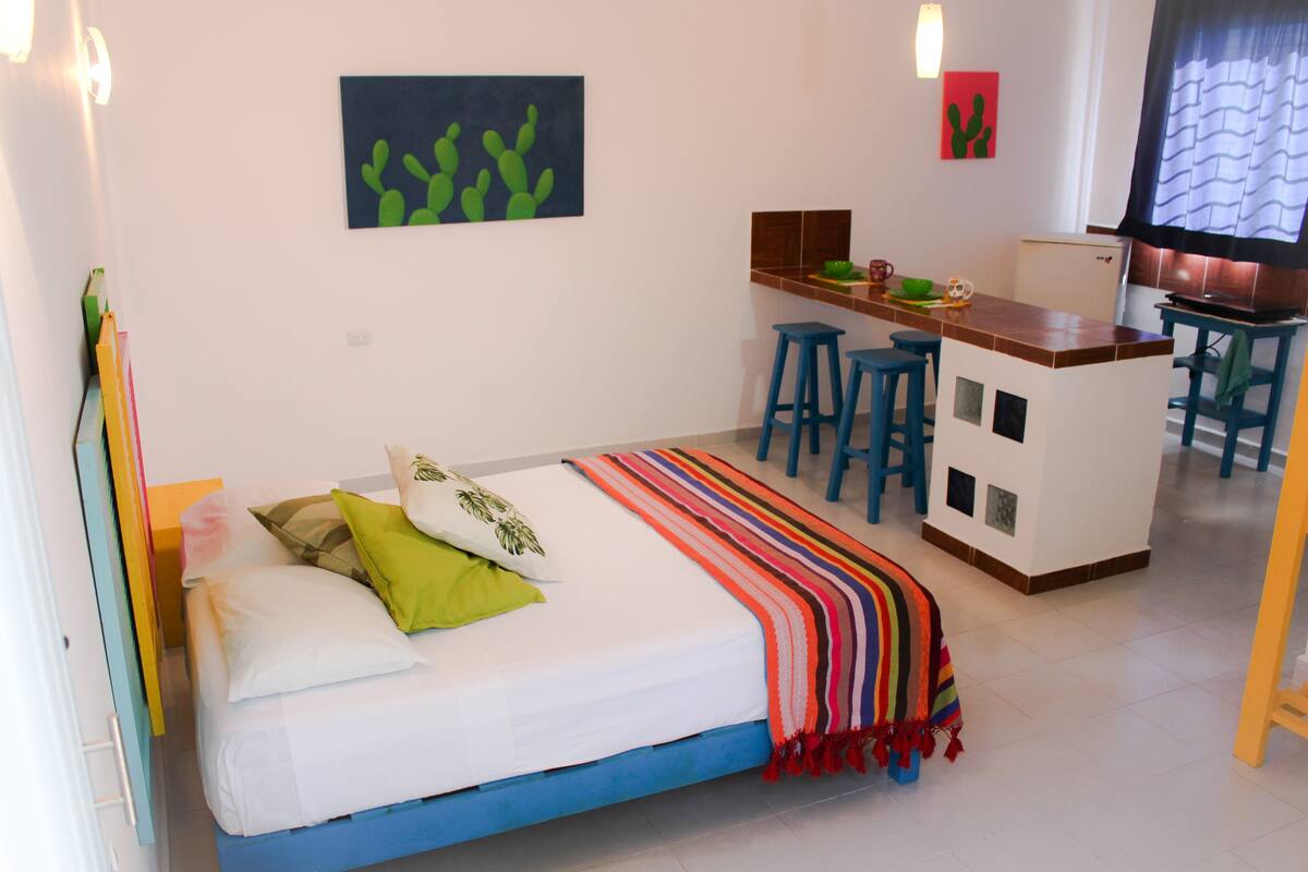 Quarto com cama, travesseiros, manta listrada colorida, escrivaninha com banquinhos e cozinha.