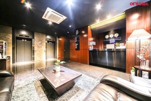 Um saguão de hotel com uma mesa de recepção, dois elevadores, uma mesinha de centro e assentos de couro.