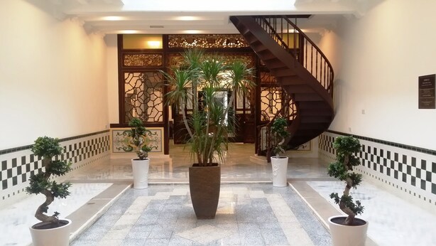 Um hall amplo com piso de mármore, uma planta em vaso central e uma escada em espiral, além de painéis decorativos nas paredes.