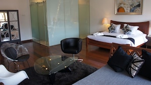 Quarto de hotel com cama, mesa de vidro, cadeira preta, sofá, banco de madeira e armário em vidro.