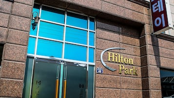 Exterior do hotel Hilton Park, com fachada de vidro transparente, uma placa de destaque e céu azul ao fundo.