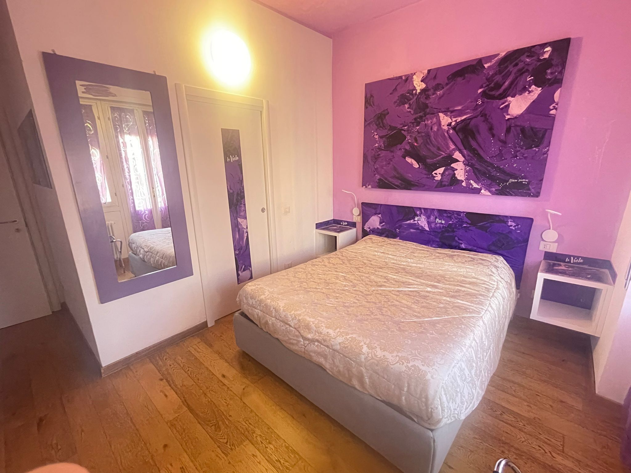 Quarto com cama, espelho, mesa de cabeceira, televisão na parede e uma porta.