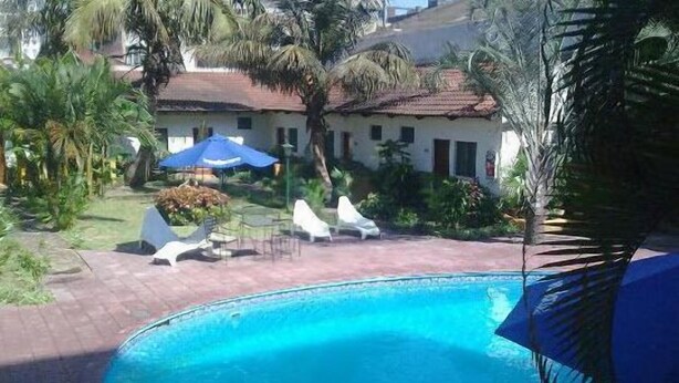 Piscina com espreguiçadeiras e guarda-sóis, cercada por palmeiras e um edifício com telhado vermelho.