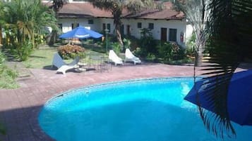 Piscina com espreguiçadeiras e guarda-sóis, cercada por palmeiras e um edifício com telhado vermelho.
