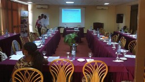 Sala de conferências com mesas grandes, cadeiras e uma tela de projetor exibindo uma apresentação.