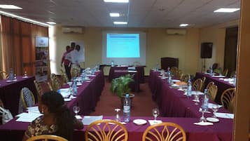 Sala de conferências com mesas grandes, cadeiras e uma tela de projetor exibindo uma apresentação.