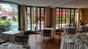 Restaurante com mesas ao ar livre, grandes janelas e vista para uma piscina e vegetação.