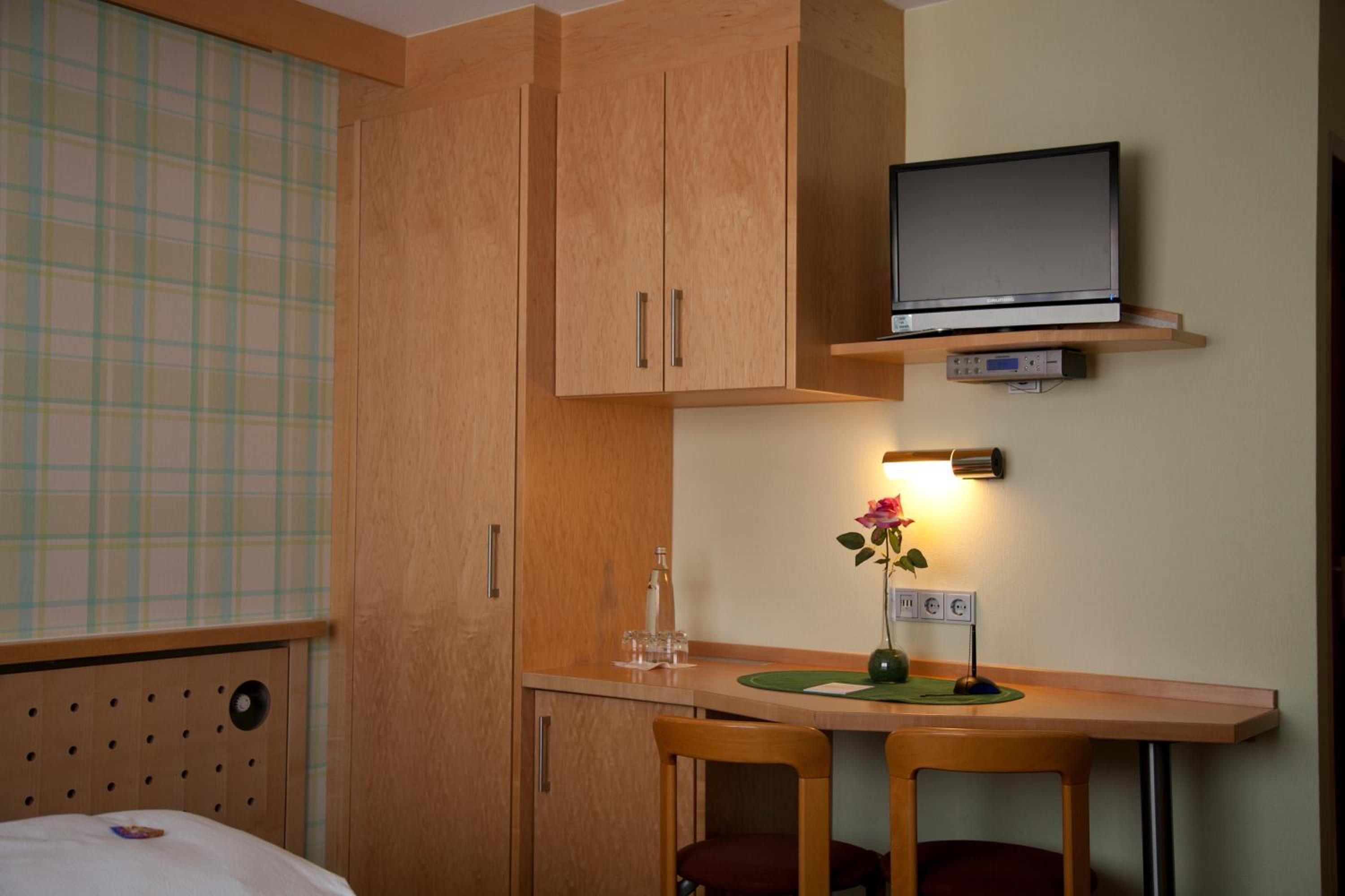 Quarto de hotel com cama, armários de madeira, uma mesa com duas cadeiras, televisão e uma pequena mesa com um vaso e uma flor.